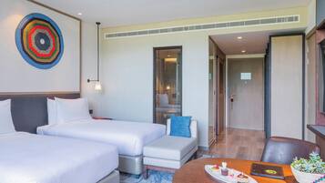 Quarto de hotel com duas camas, um espaço de estar pequeno, uma mesa de jantar com um laptop e uma tigela decorativa, além de uma obra de arte colorida na parede.