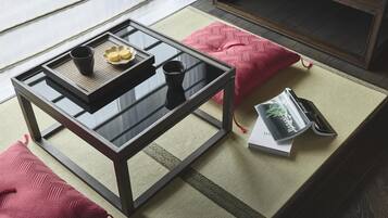 Quarto com piso de tatame, uma mesa de vidro com um tabuleiro, uma revista e um almofada vermelha.