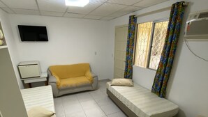 Quarto compacto com sofá, duas camas de solteiro, pequena mesa de trabalho com computador e televisão na parede.