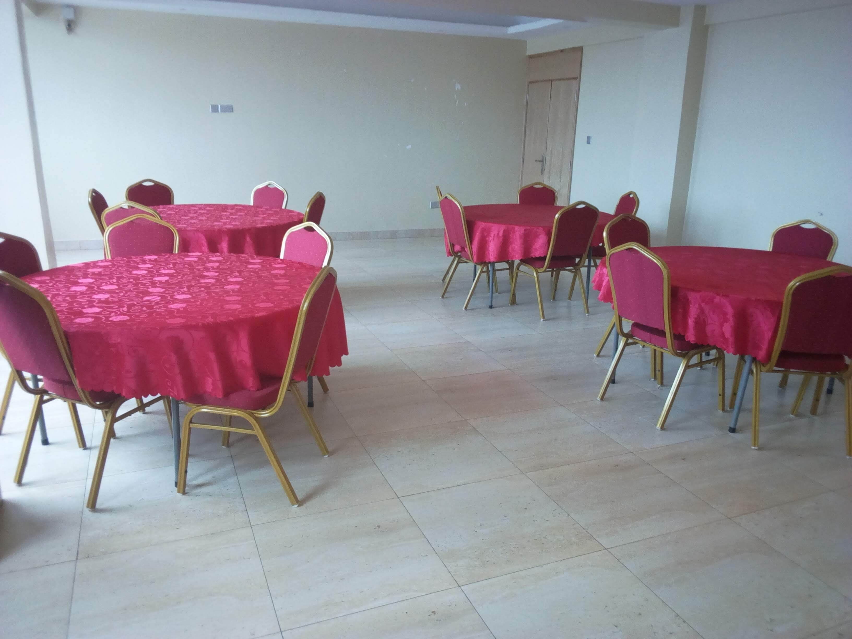 Uma sala de conferências com várias mesas redondas cobertas por toalhas vermelhas, cada uma rodeada por cadeiras vermelhas com estrutura dourada. O chão é ladrilhado e as paredes são simples.
