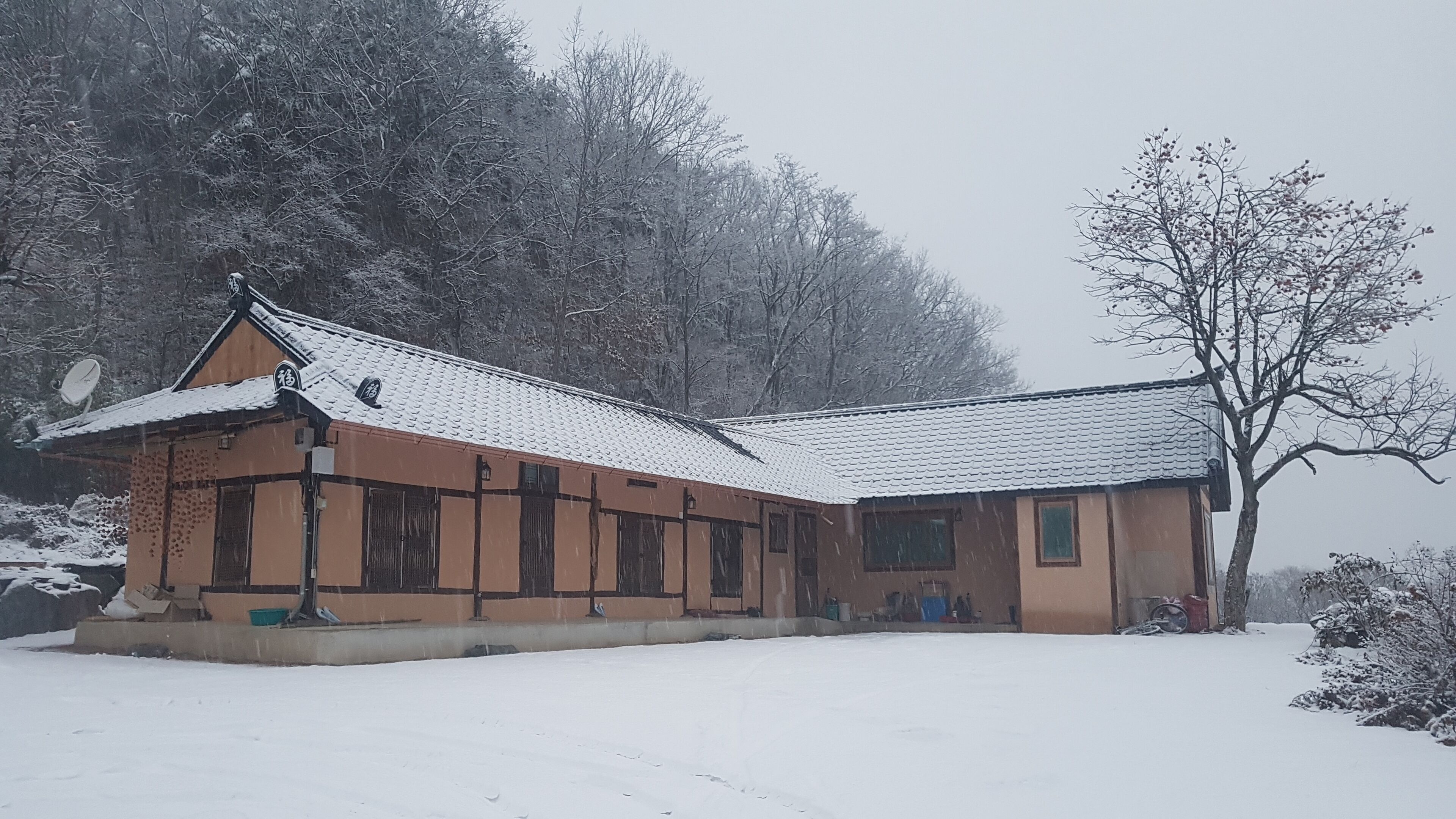 Um edifício tradicional com telhado de telhas e antena parabólica, rodeado por terreno coberto de neve e árvores.
