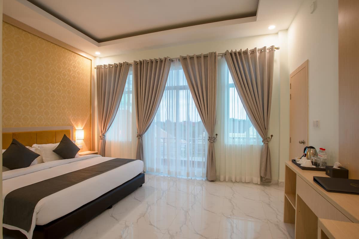 Quarto de hotel com cama, escrivaninha e uma janela grande com cortinas.