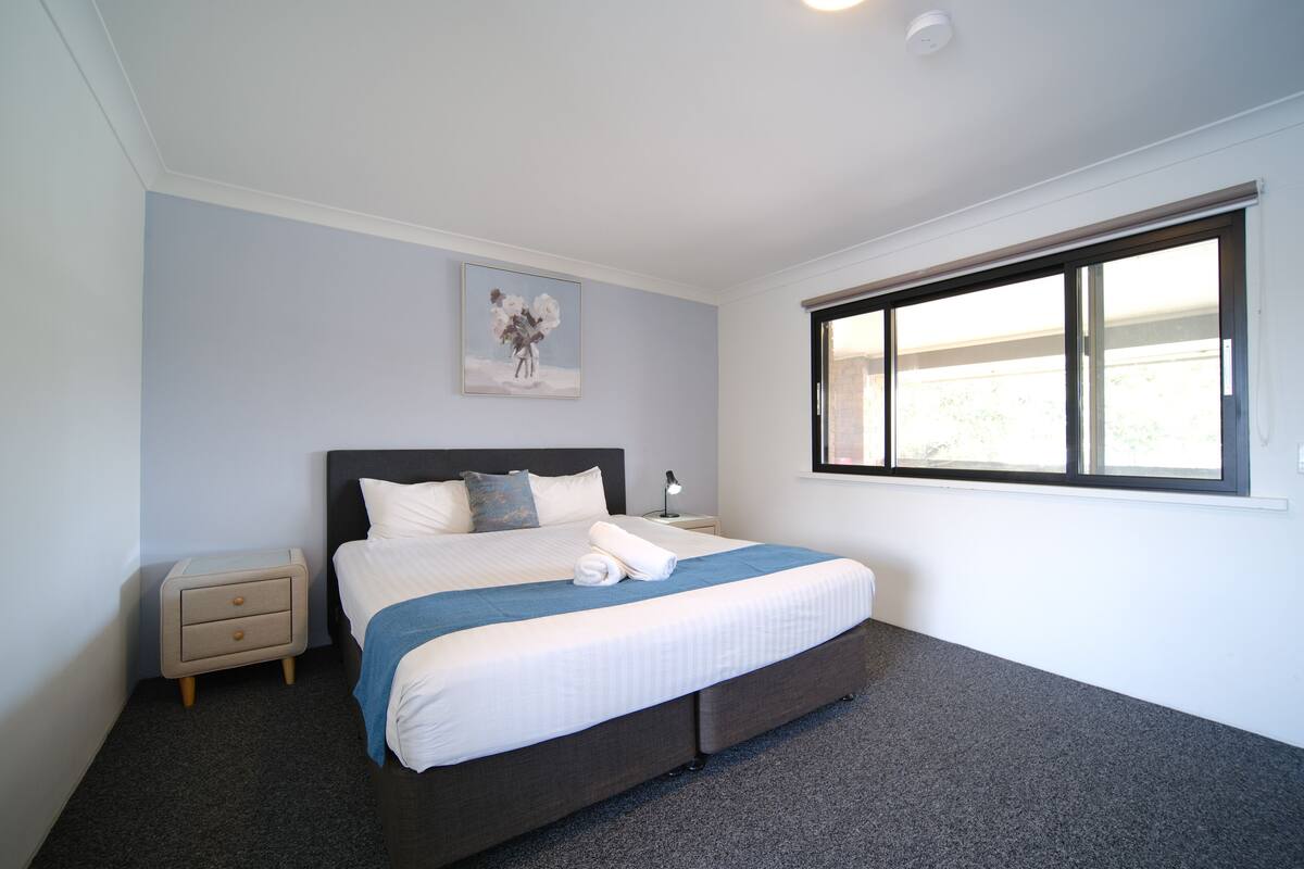 Um quarto com cama, mesa de cabeceira, abajur, janela e um quadro na parede.