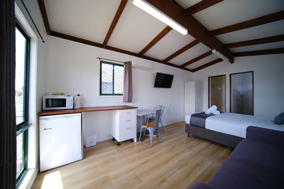 Quarto compacto com cozinha compacta, uma cama, escrivaninha e uma cadeira.