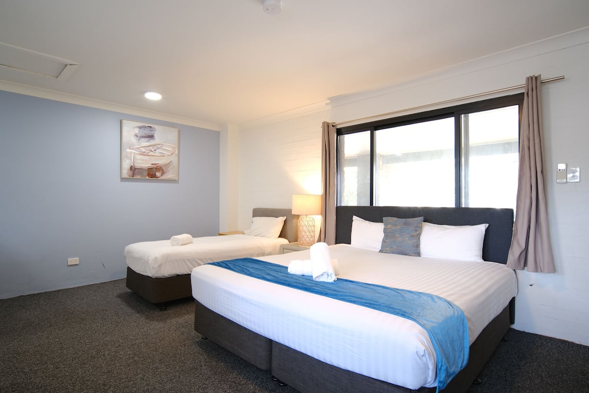 Quarto de hotel com duas camas, uma janela grande com cortinas, um quadro pendurado na parede e um abajur.
