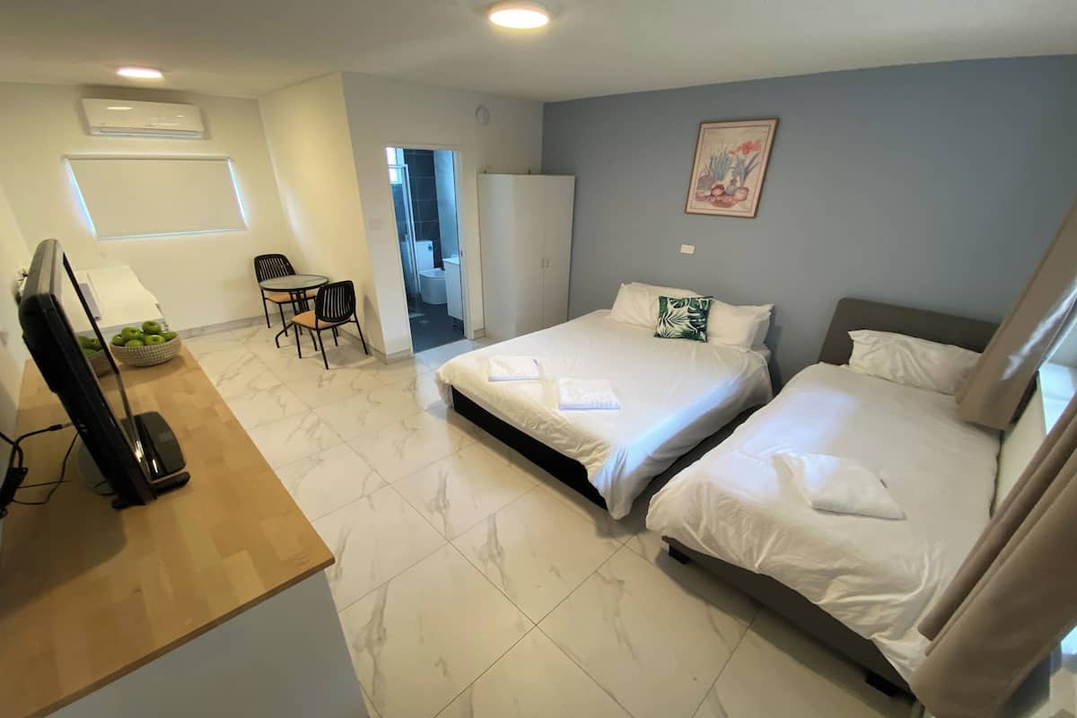 Quarto de hotel com duas camas, uma escrivaninha com um computador e uma luminária, uma cadeira e um banheiro visível por uma porta aberta.