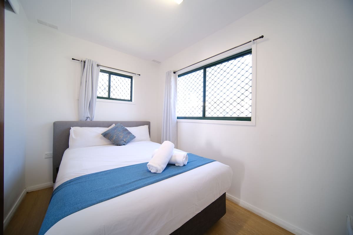 Uma cama bem arrumada com roupa de cama branca e azul, uma cabeceira cinza e duas janelas com cortinas.
