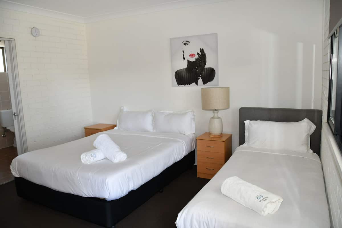 Quarto de hotel com duas camas, roupa de cama branca, um quadro na parede, uma luminária e um relógio.