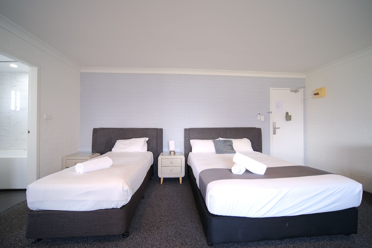 Quarto de hotel com duas camas, uma pequena mesa de cabeceira com abajur e porta para o banheiro.
