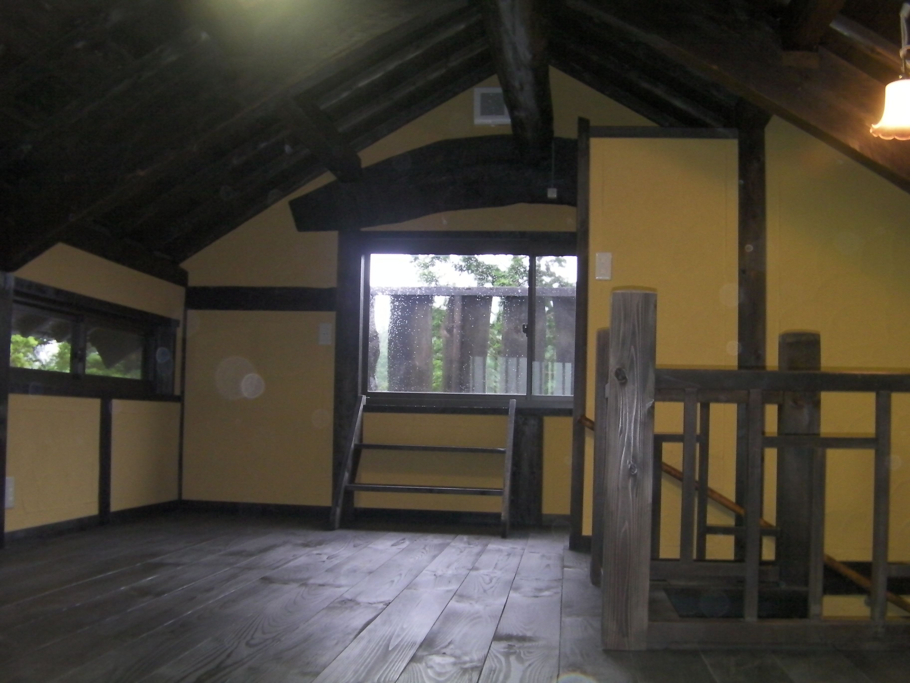 Quarto de sótão com piso de madeira, janela com vista para vegetação e um corrimão de madeira.