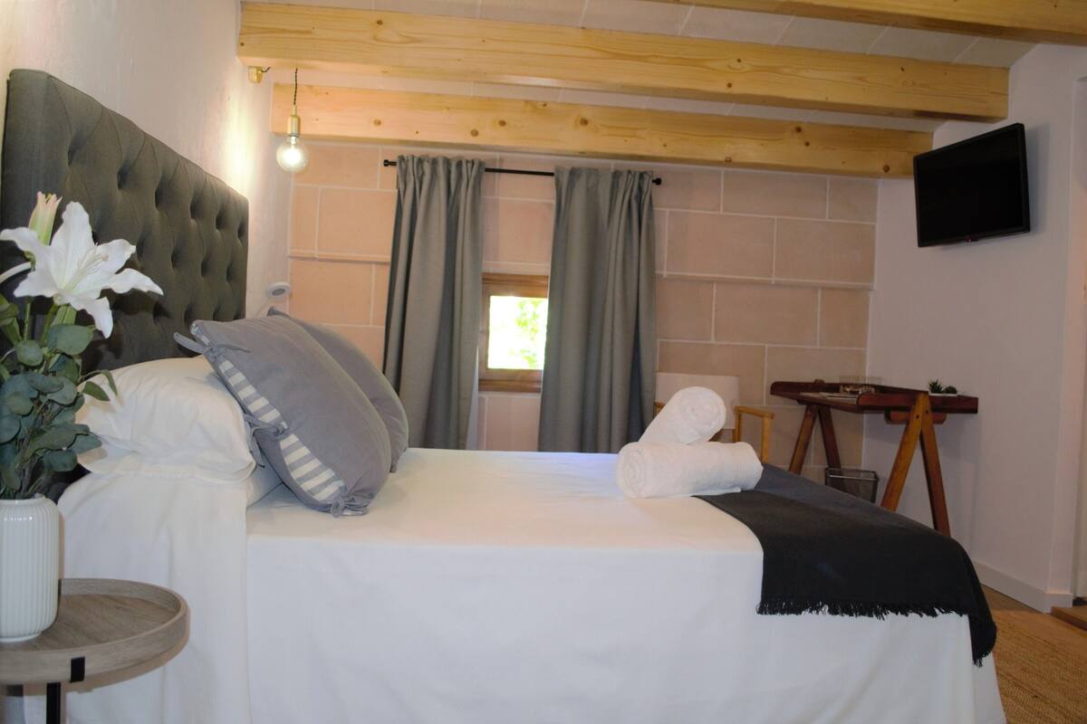 Quarto com cabeceira estofada, cama com roupa de cama branca, almofadas cinzas, manta preta, mesa de cabeceira de madeira com um vaso de flores e uma cadeira de madeira.