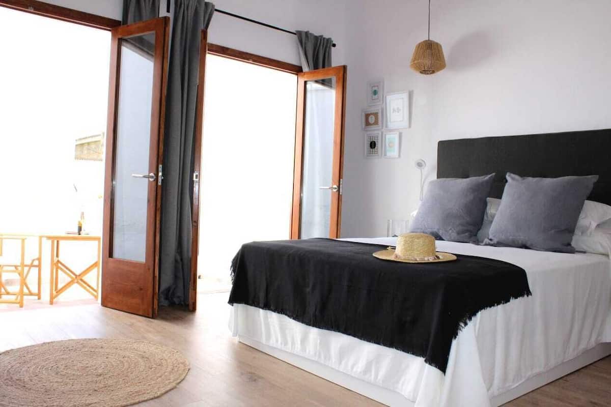 Quarto com cama, mesa de cabeceira, luminária pendente e vista por uma porta aberta.