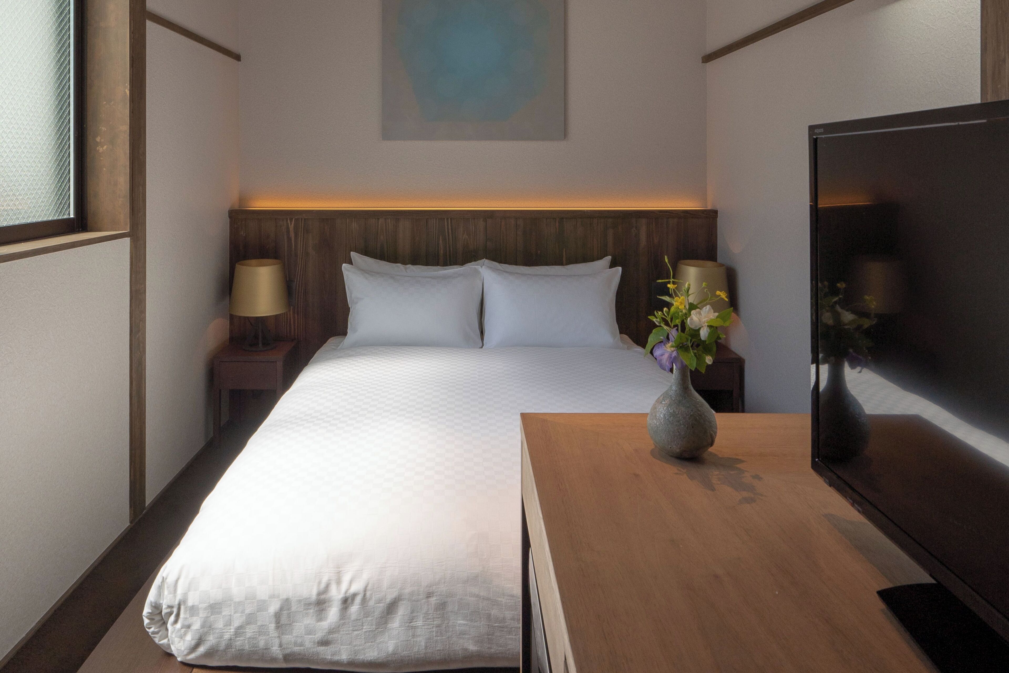 Quarto de hotel com uma cama grande, uma mesinha de cabeceira de madeira com luminárias, uma TV e um vaso com flores sobre uma mesa de madeira.