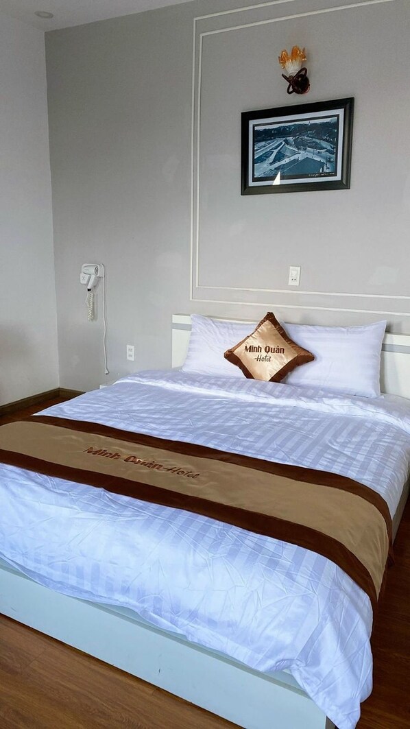 Uma cama bem arrumada com edredom branco e marrom, um travesseiro com a inscrição "Minh Quận Hotel" e um quadro na parede.