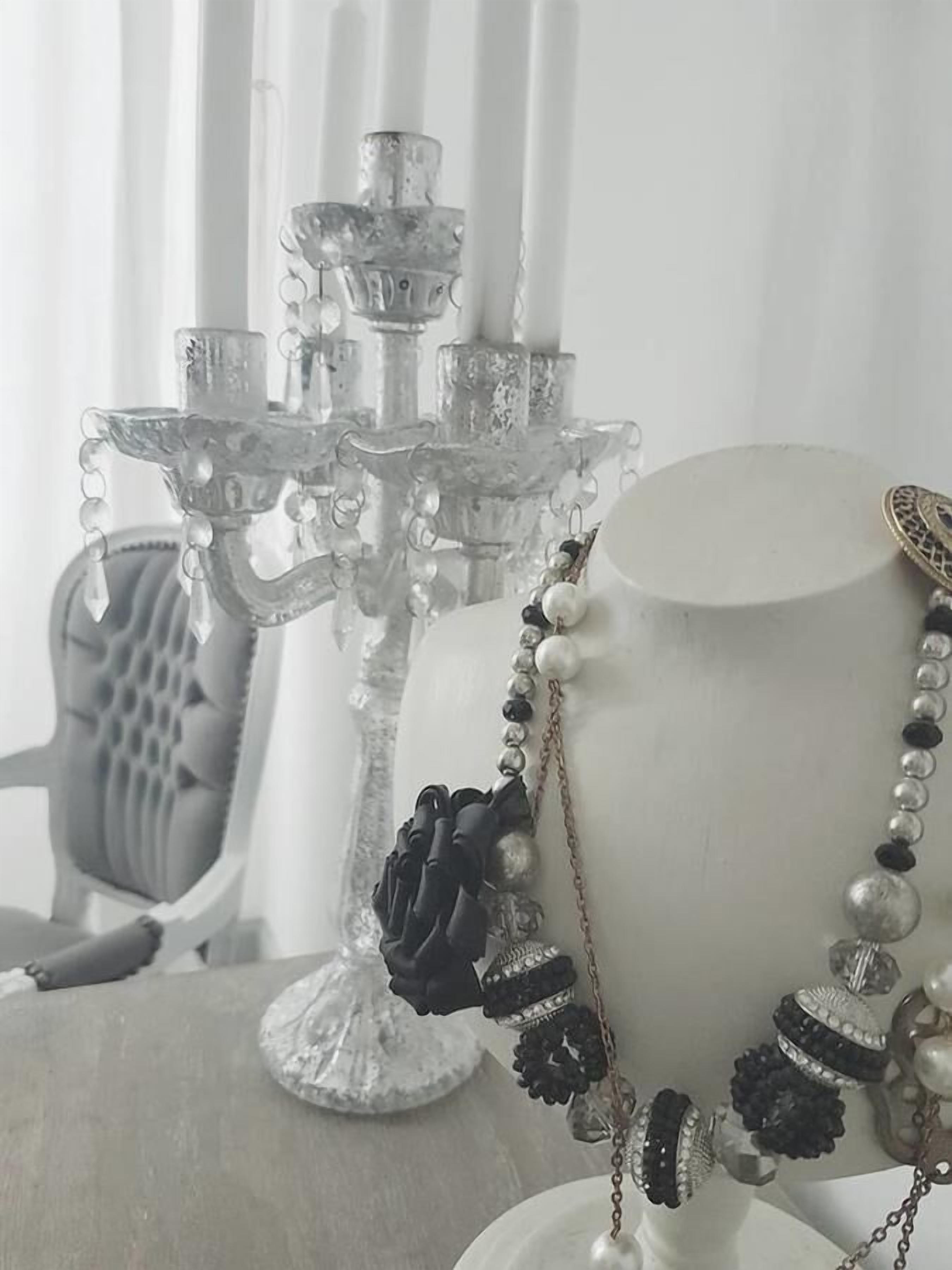 Um manequim com um colar, um candelabro de vidro e uma cadeira em um ambiente com fundo branco.