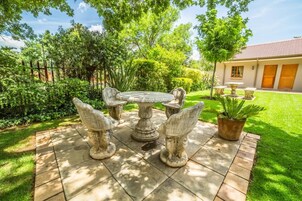 Um pátio com uma mesa e cadeiras de pedra, rodeado por vegetação exuberante e uma casa ao fundo.
