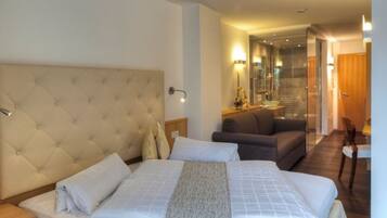 Quarto de hotel com uma cama grande, um sofá, uma mesa de jantar e um banheiro visível através de uma porta de vidro.