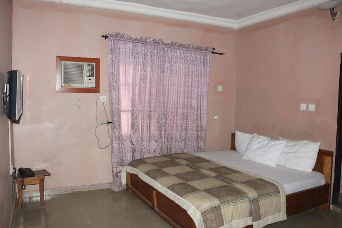 Quarto com cama, ar-condicionado fixado na parede, cortina e uma mesinha com um telefone.