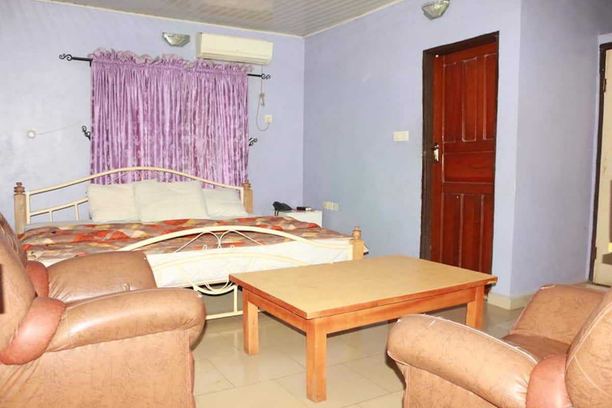 Quarto com cama, mesa de madeira, duas poltronas de couro, janela com cortinas e porta de madeira.