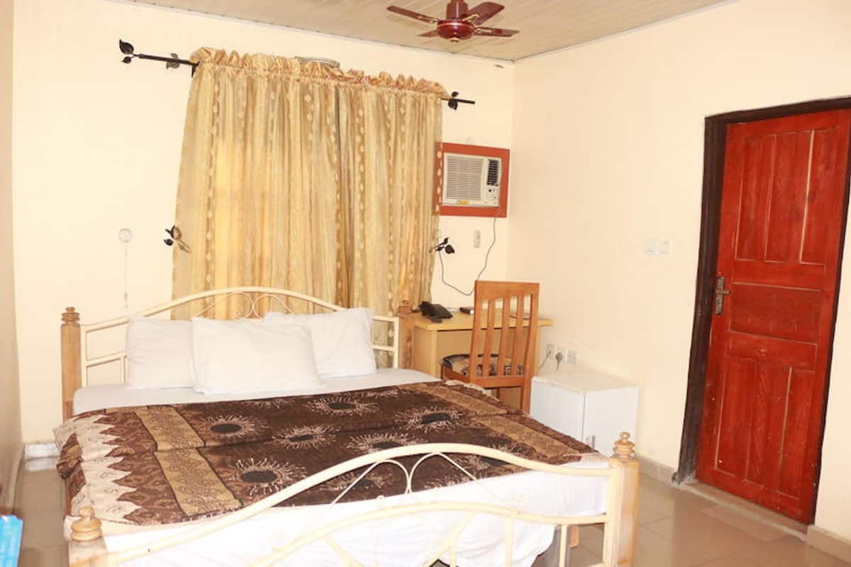 Quarto com cama, escrivaninha, cadeira, porta vermelha e ventoinha no teto.