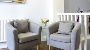 Duas poltronas cinzas com pernas de madeira, um radiador branco, um vaso de vidro com flores e um corrimão branco.