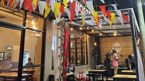 Um restaurante com mesas ao ar livre, uma placa com a inscrição 'NYOR!' e uma decoração festiva com bandeirinhas triangulares.