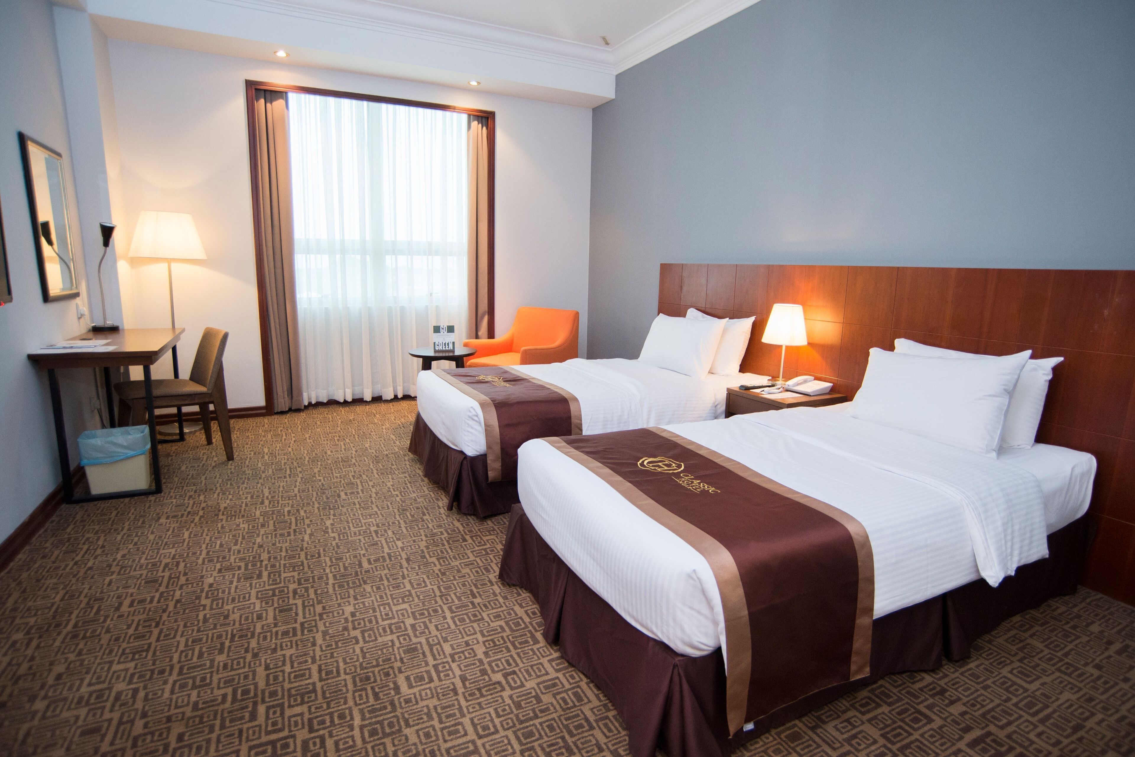 Quarto de hotel com duas camas, uma escrivaninha, uma cadeira e uma janela grande com cortinas.