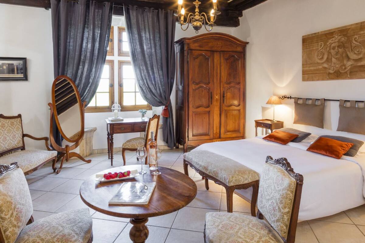 Quarto de hotel com cama, guarda-roupa de madeira, secretária com cadeira, mesa redonda com cadeiras, espelho e lustre.