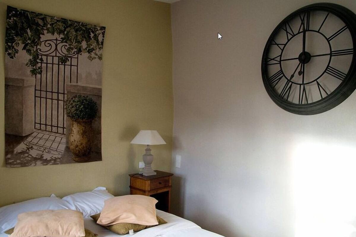 Quarto de hotel com cama, mesa de cabeceira com luminária, relógio de parede grande e pintura de um portão.