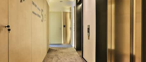 Corredor de hotel com carpete estampado, uma porta com número e um elevador.