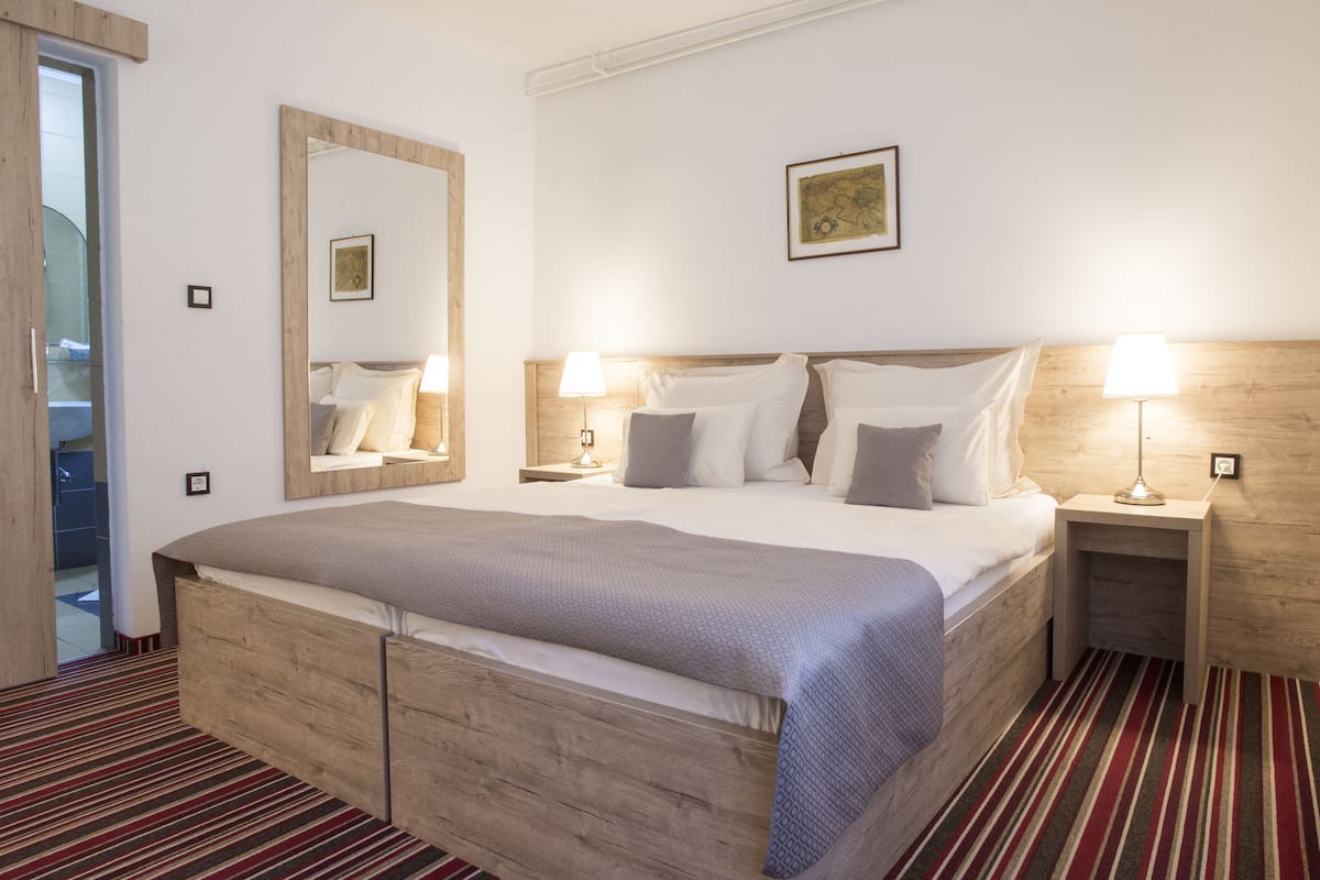 Uma cama bem arrumada com cabeceira de madeira, duas mesas de cabeceira com abajures, um espelho grande e um tapete listrado em vermelho e branco.