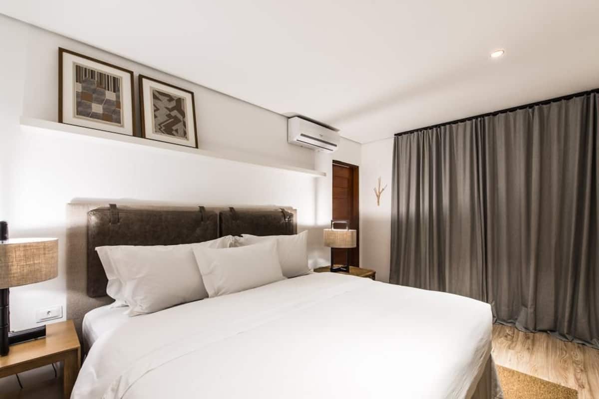 Quarto de hotel com uma cama grande, mesa de cabeceira, luminária, ar condicionado e janela com cortinas.