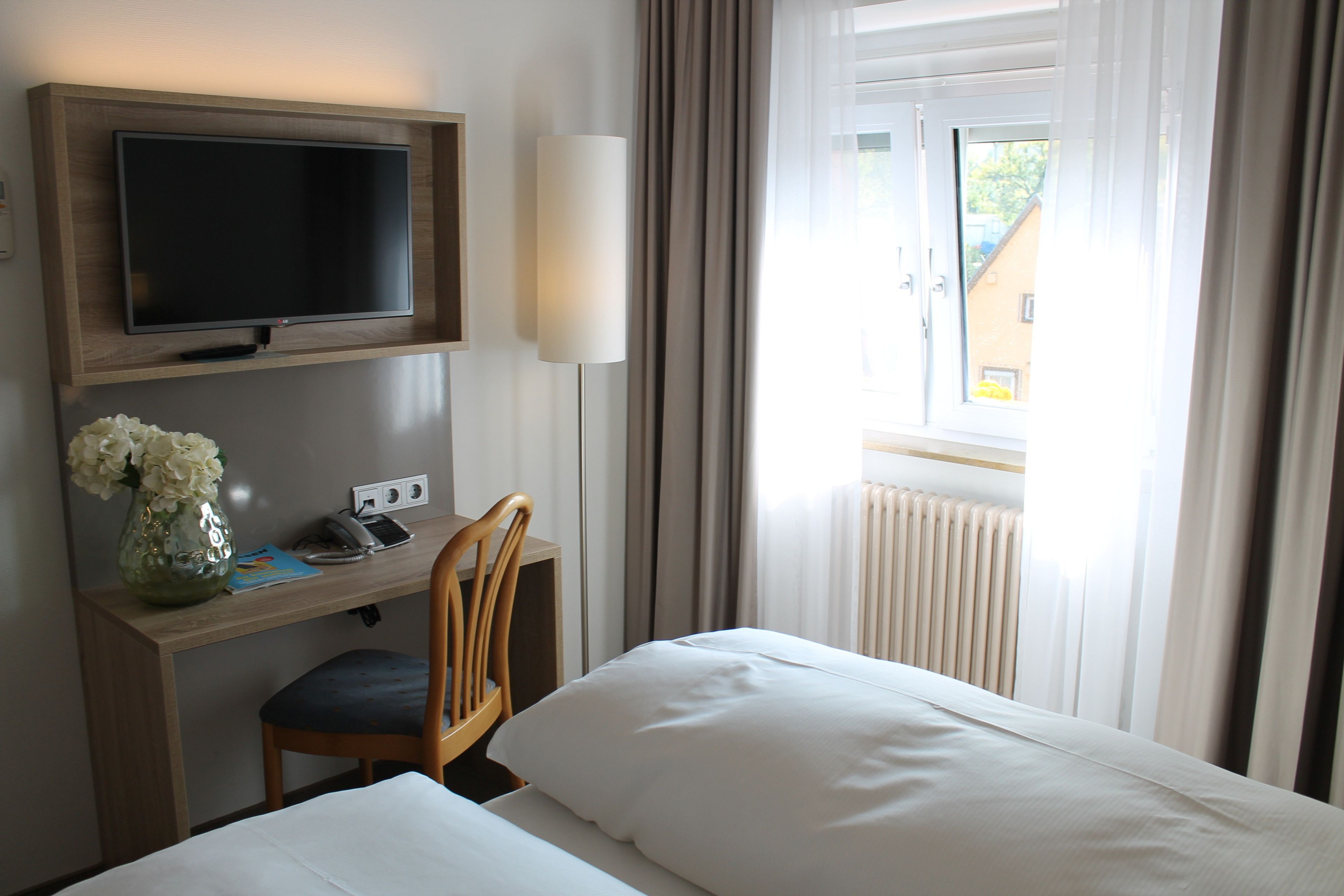 Quarto de hotel com uma cama, uma mesa com cadeira, televisão e janela com cortinas.