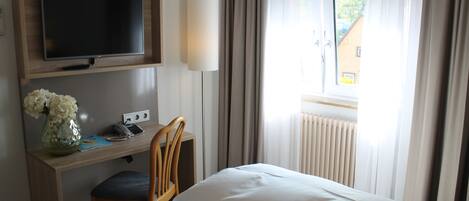 Quarto de hotel com uma cama, uma mesa com cadeira, televisão e janela com cortinas.