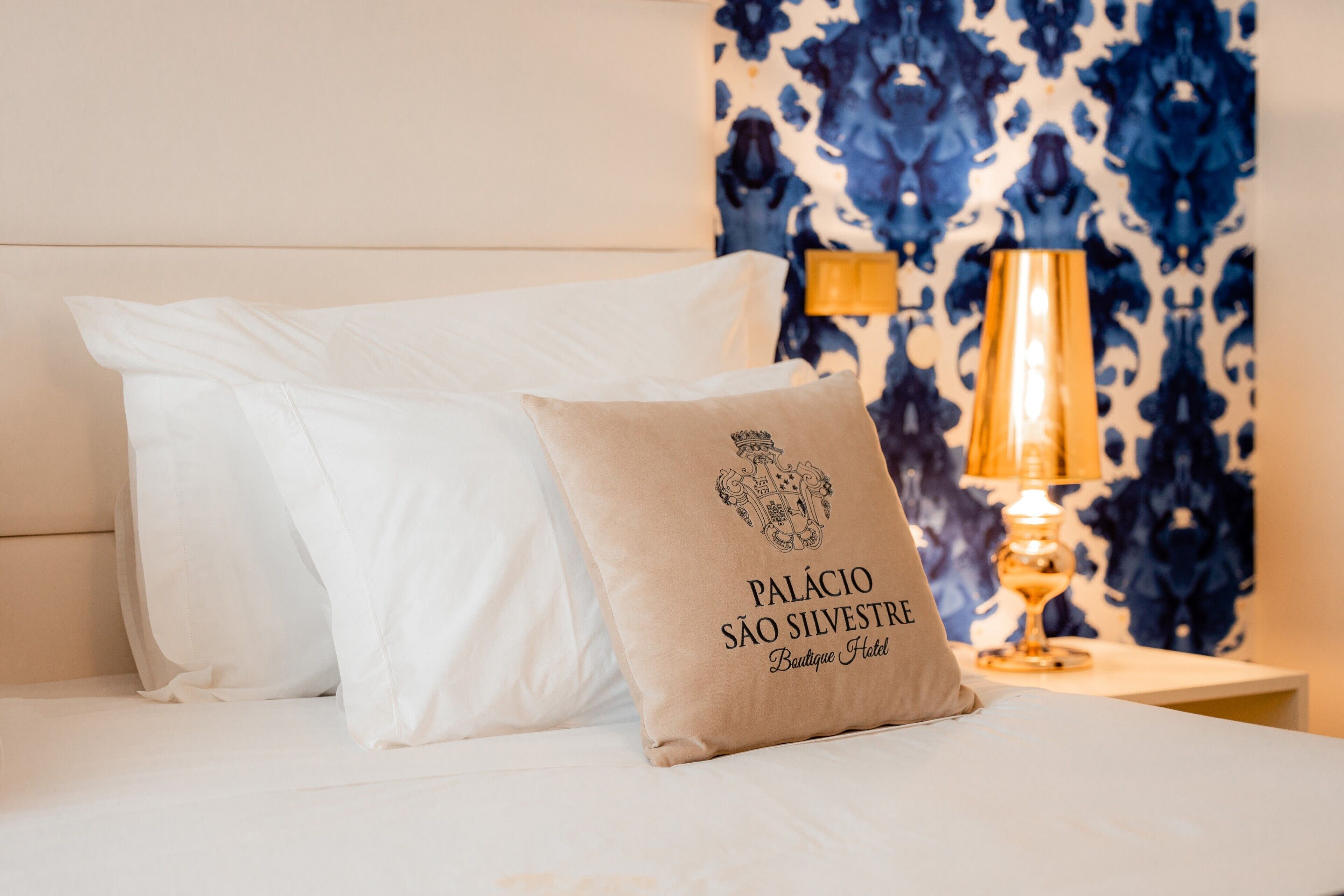 Cama com roupa de cama branca e travesseiro com a inscrição "Palácio São Silvestre Boutique Hotel".