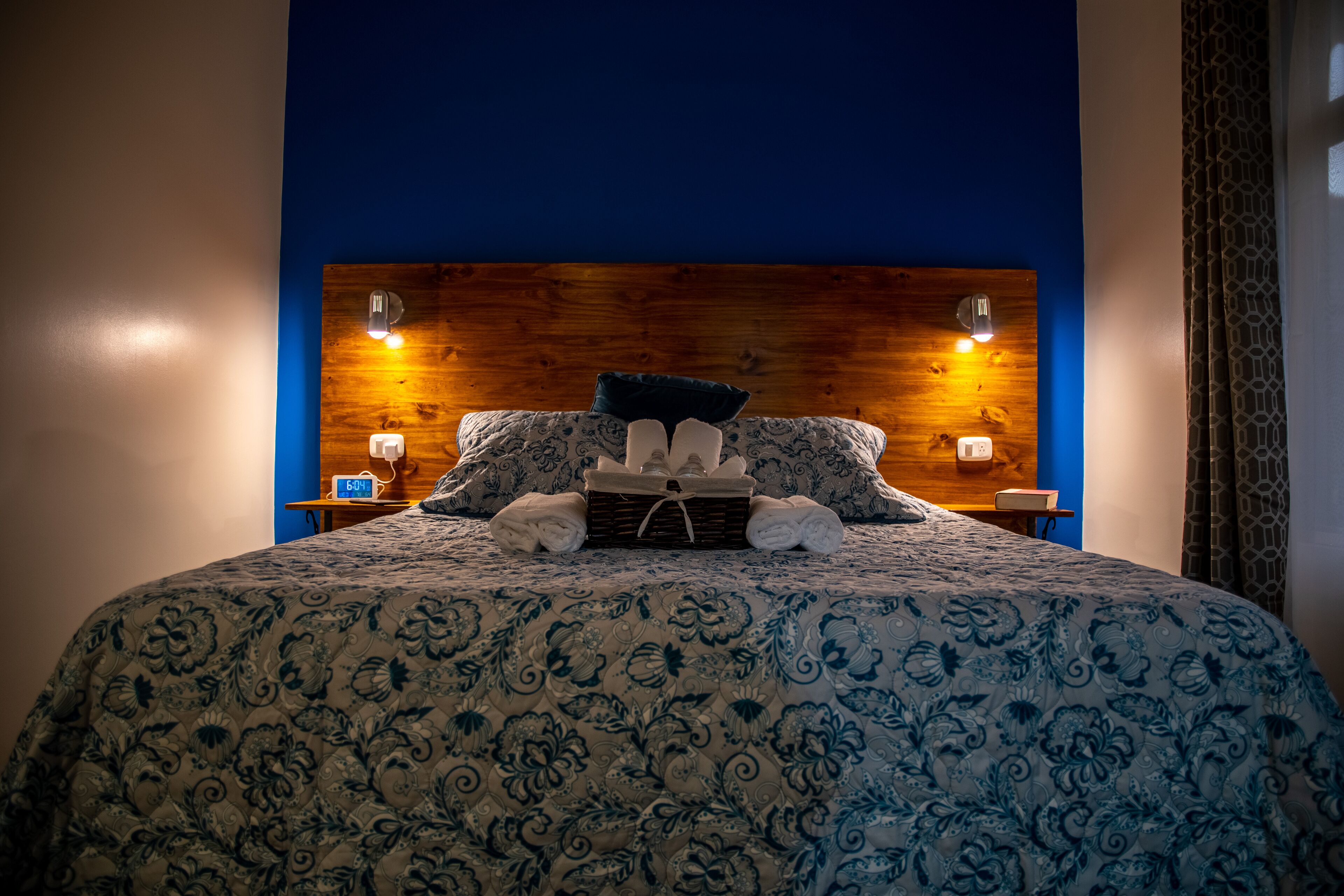 Uma cama bem arrumada com um edredom estampado com motivos florais, uma mesa de cabeceira com abajur e uma cabeceira de madeira.