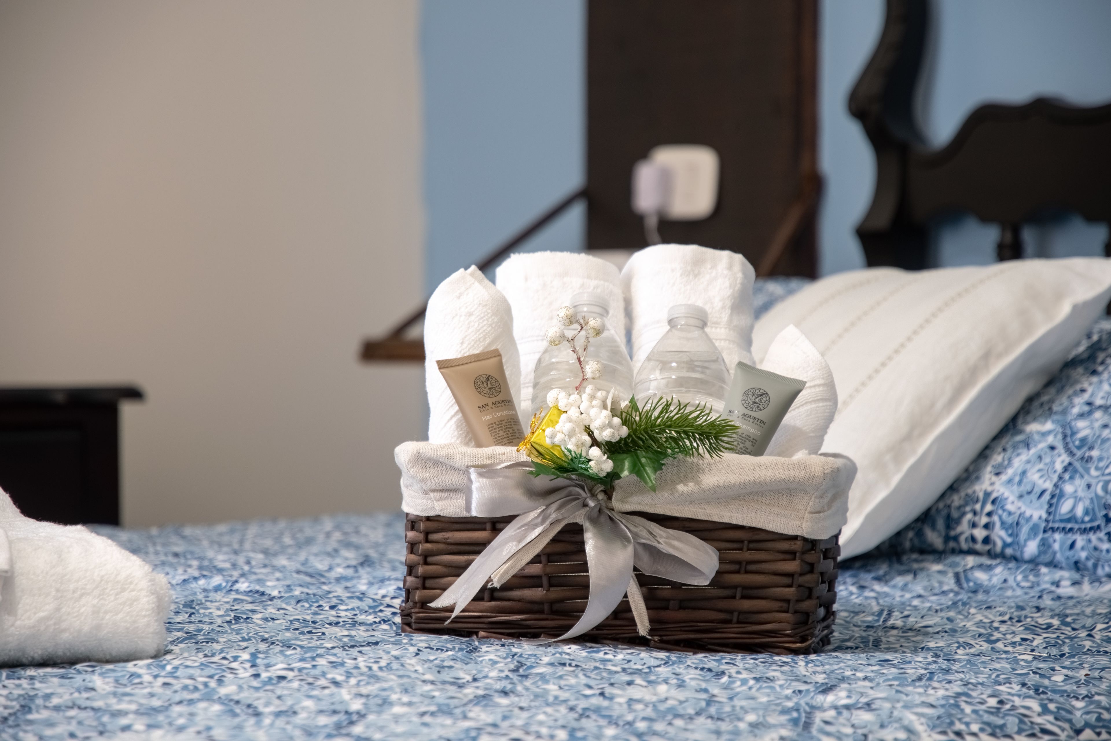 Uma cesta de vime com produtos de higiene e toalhas sobre uma cama com colcha azul estampada.