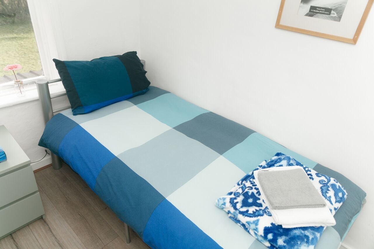 Uma cama bem arrumada com colcha xadrez azul e cinza, travesseiro estampado em azul e branco, e um quadro na parede.