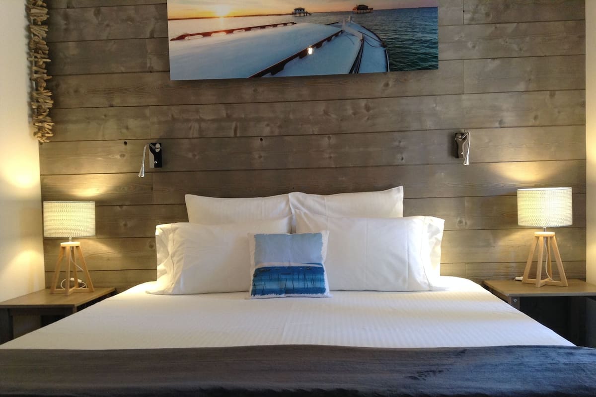 Quarto de hotel com cabeceira de madeira, cama grande com roupa de cama branca, duas luminárias de cabeceira e uma pintura de um pôr do sol sobre a água.
