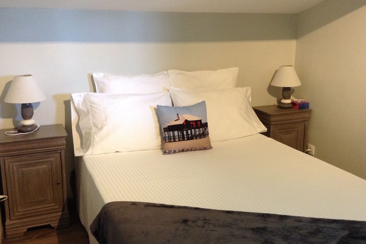 Cama bem arrumada com lençóis brancos e uma única almofada decorativa. Duas mesas de cabeceira, cada uma com uma luminária e uma pequena caixa. O quarto tem um design simples e clean, com paredes em tons claros.