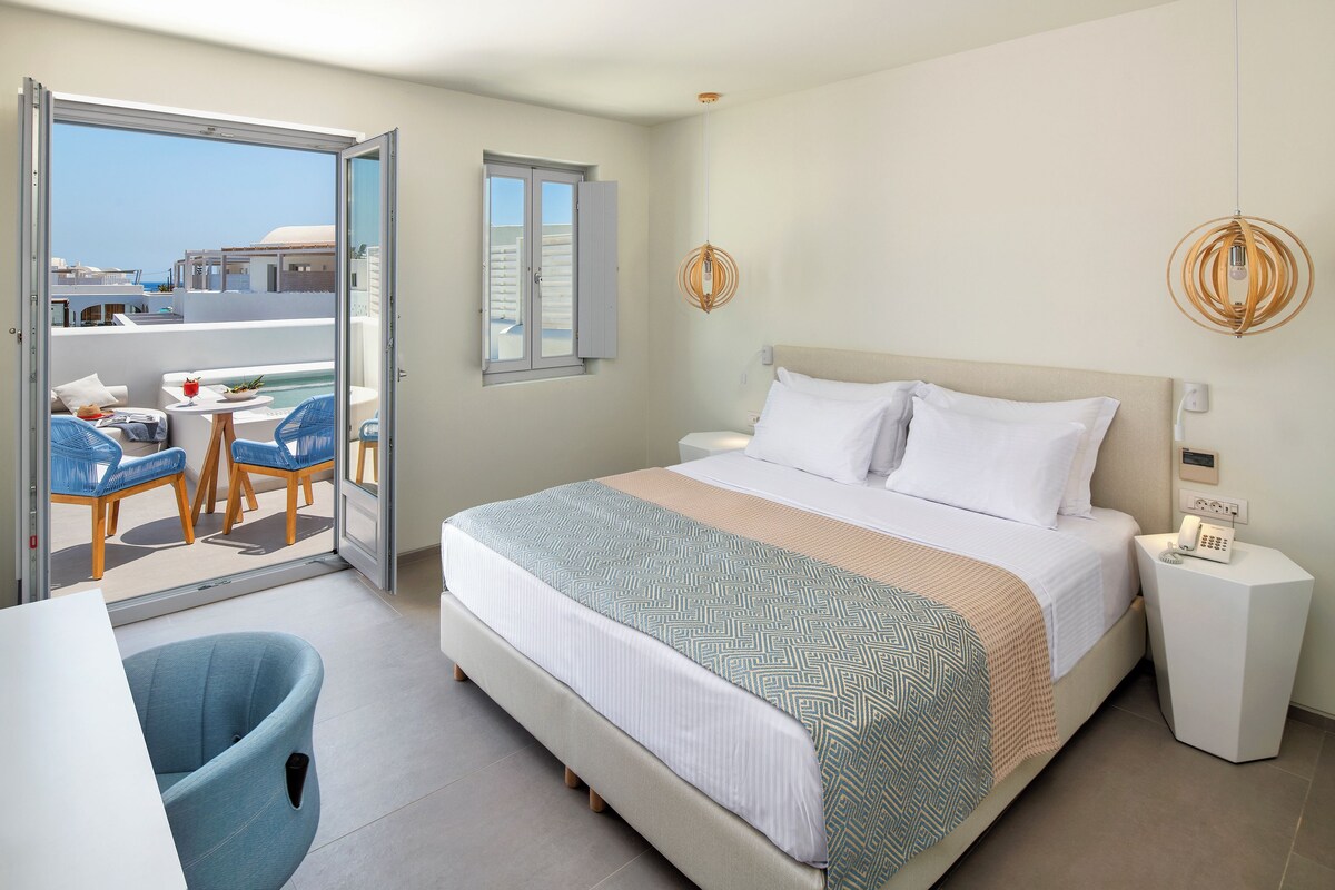 Quarto de hotel moderno com cama, escrivaninha e varanda com cadeiras.