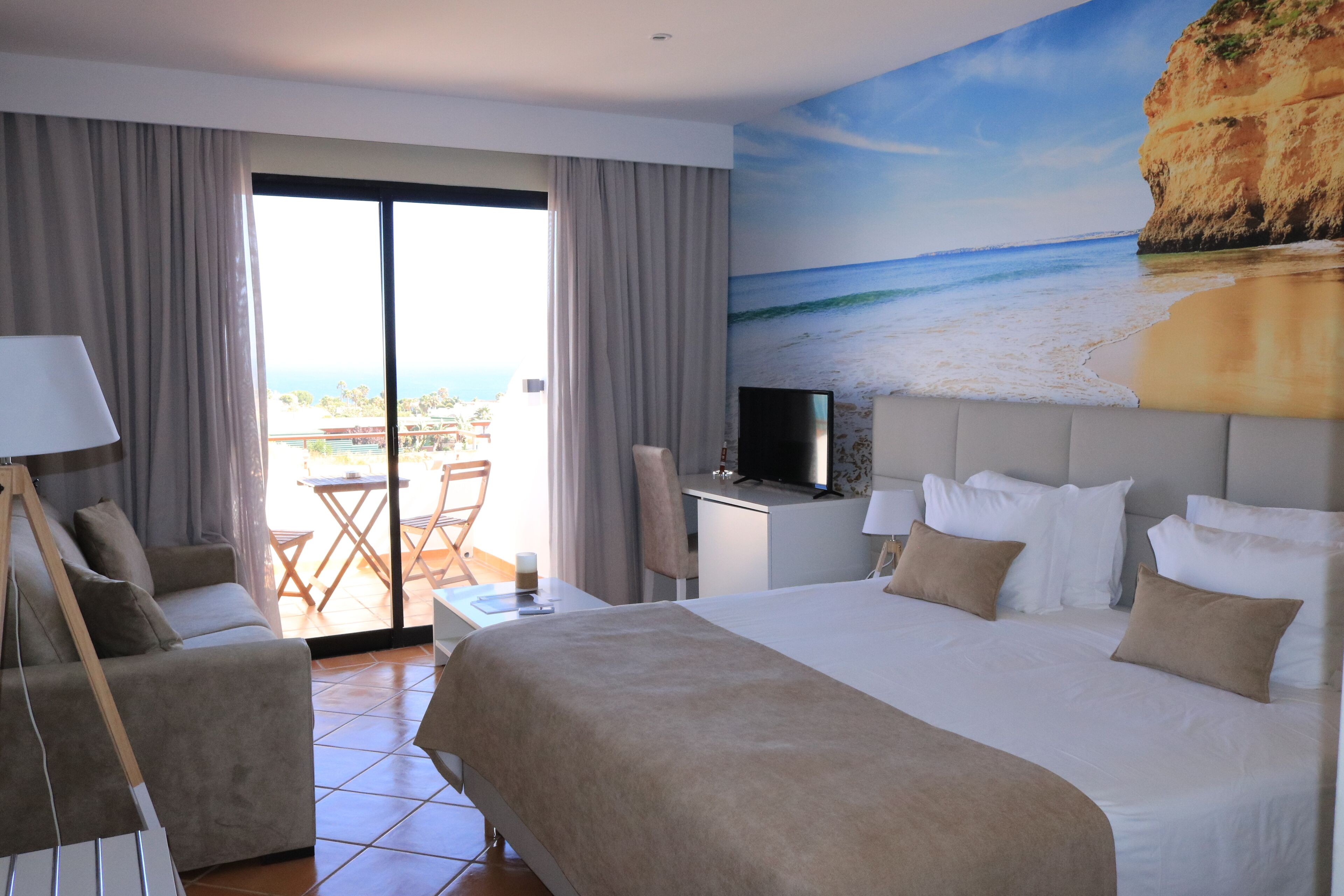 Quarto de hotel com cama, escrivaninha, televisão, cadeira e vista para a praia através de uma grande janela.