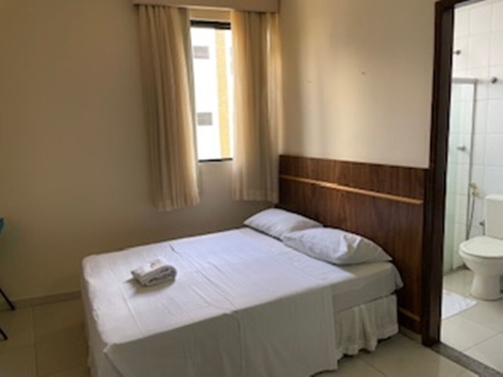 Quarto de hotel com cama, janela com cortinas e vista para um banheiro.