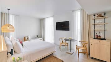 Quarto de hotel com cama, televisão, pequena mesa com cadeiras, armário com garrafas e prateleira decorativa.
