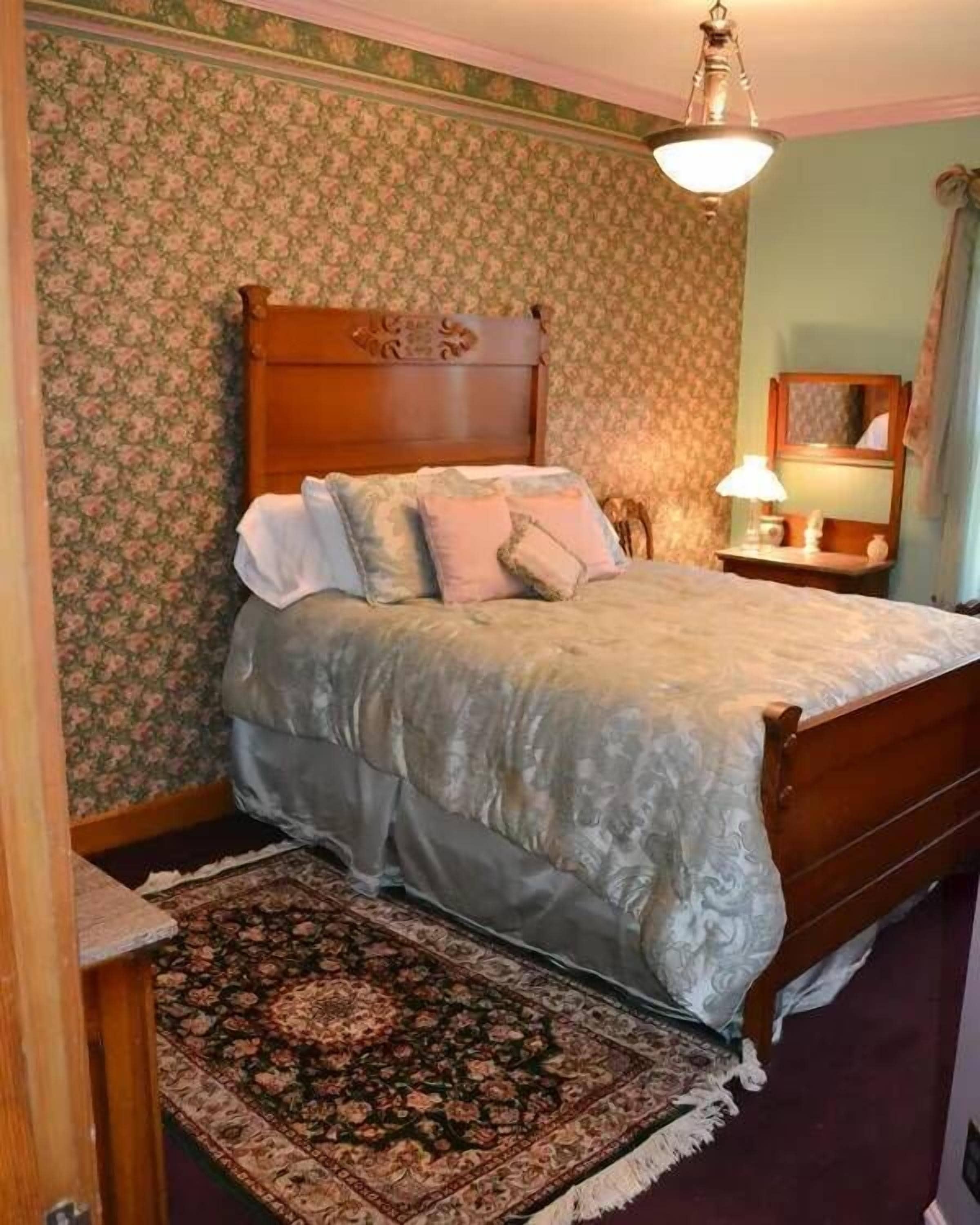 Quarto com cama de madeira, papel de parede floral, mesa de cabeceira com abajur e tapete estampado.