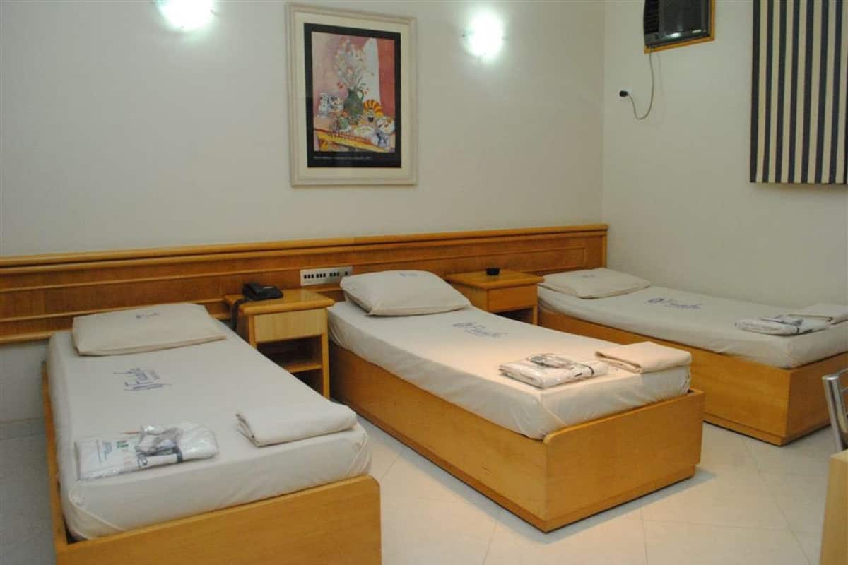 Quarto de hotel com três camas de madeira, uma mesa de cabeceira com abajur, um quadro na parede e uma janela com persianas.
