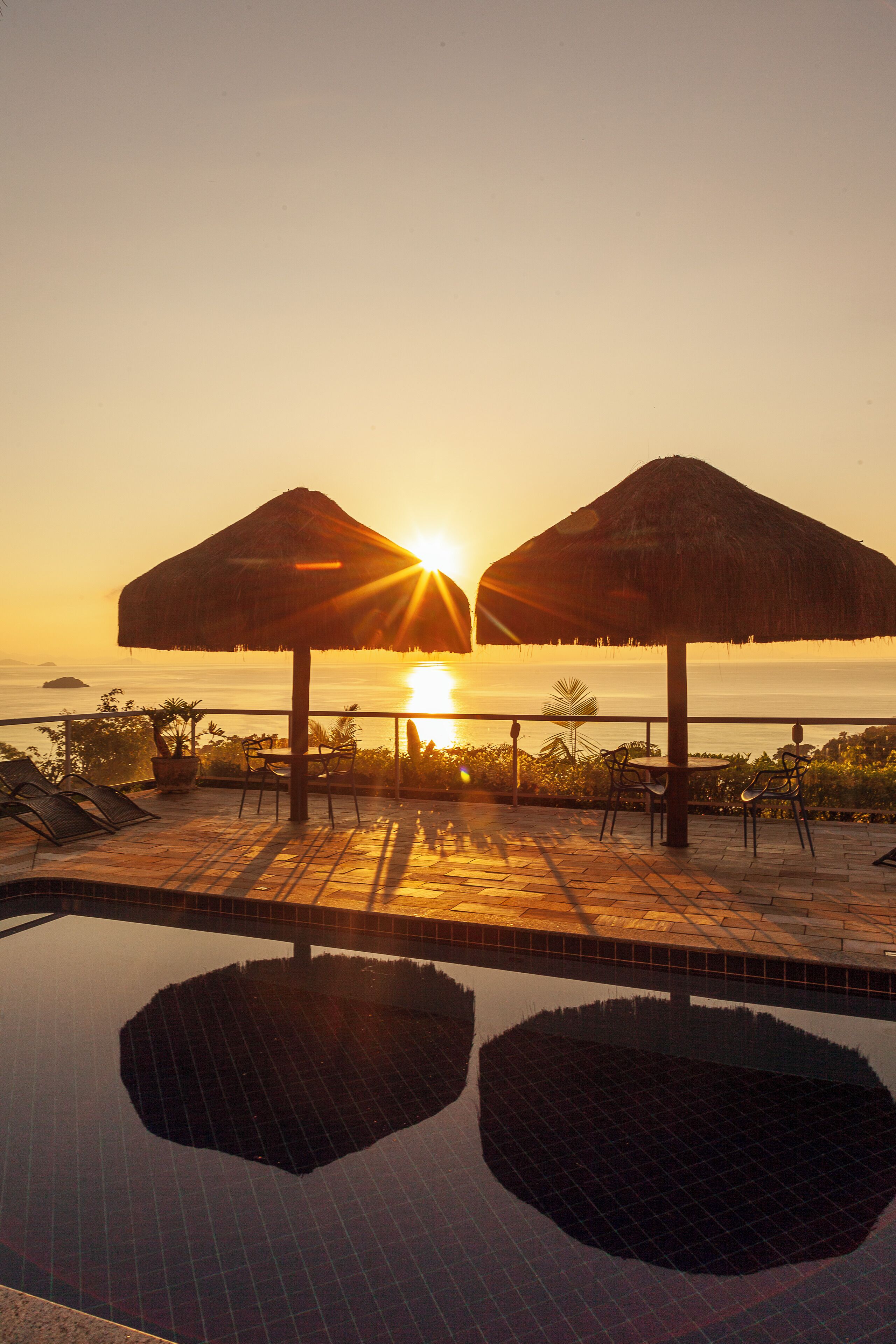 Uma área à beira da piscina com duas sombrinhas de palha, espreguiçadeiras e vista para o pôr do sol sobre o oceano.