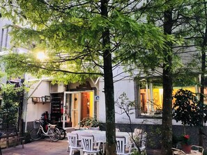 Uma área de estar externa com mesas, cadeiras, uma bicicleta e um edifício com porta e janelas.