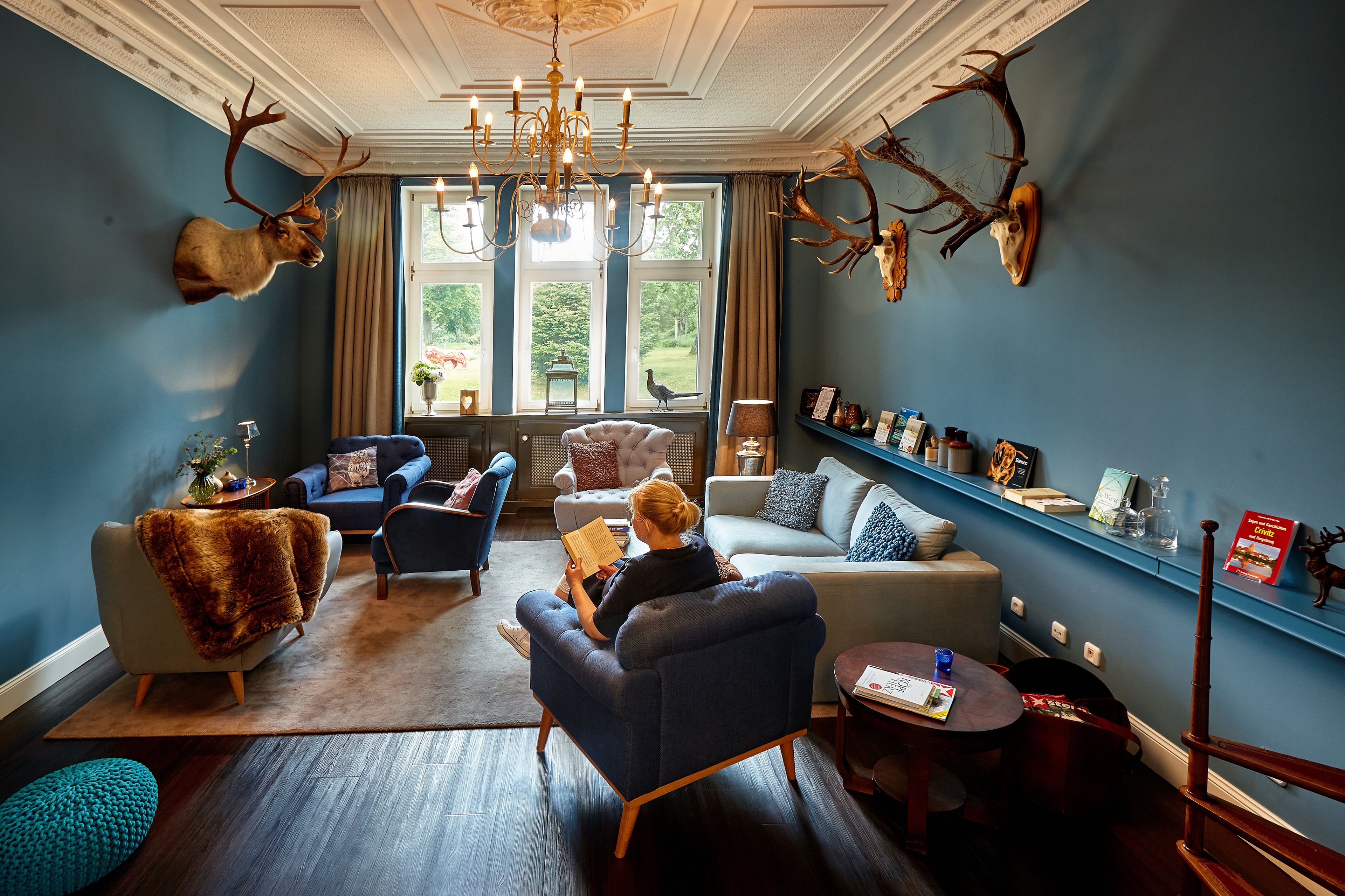 Quarto com parede azul, chifres de veado na parede, um candelabro e uma pessoa lendo um livro.