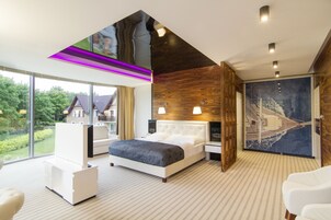 Quarto moderno com uma cama grande, uma poltrona branca e vista para uma casa através de amplas janelas.